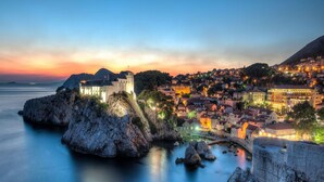Uma vila costeira com um castelo histórico numa rocha, ligada por uma ponte de pedra a uma área densamente construída com edifícios iluminados.