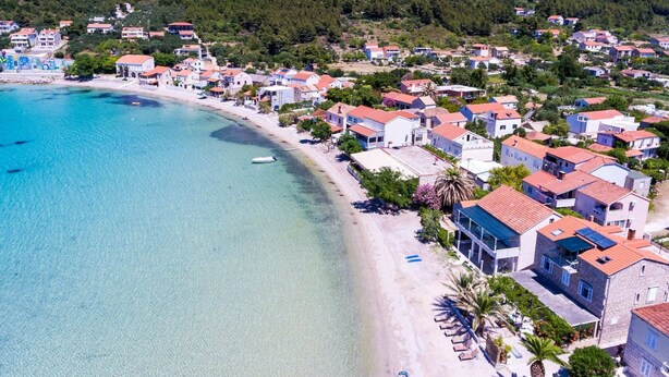 Uma vila costeira com casas ao longo da praia, águas turquesa cristalinas e colinas verdes ao fundo.