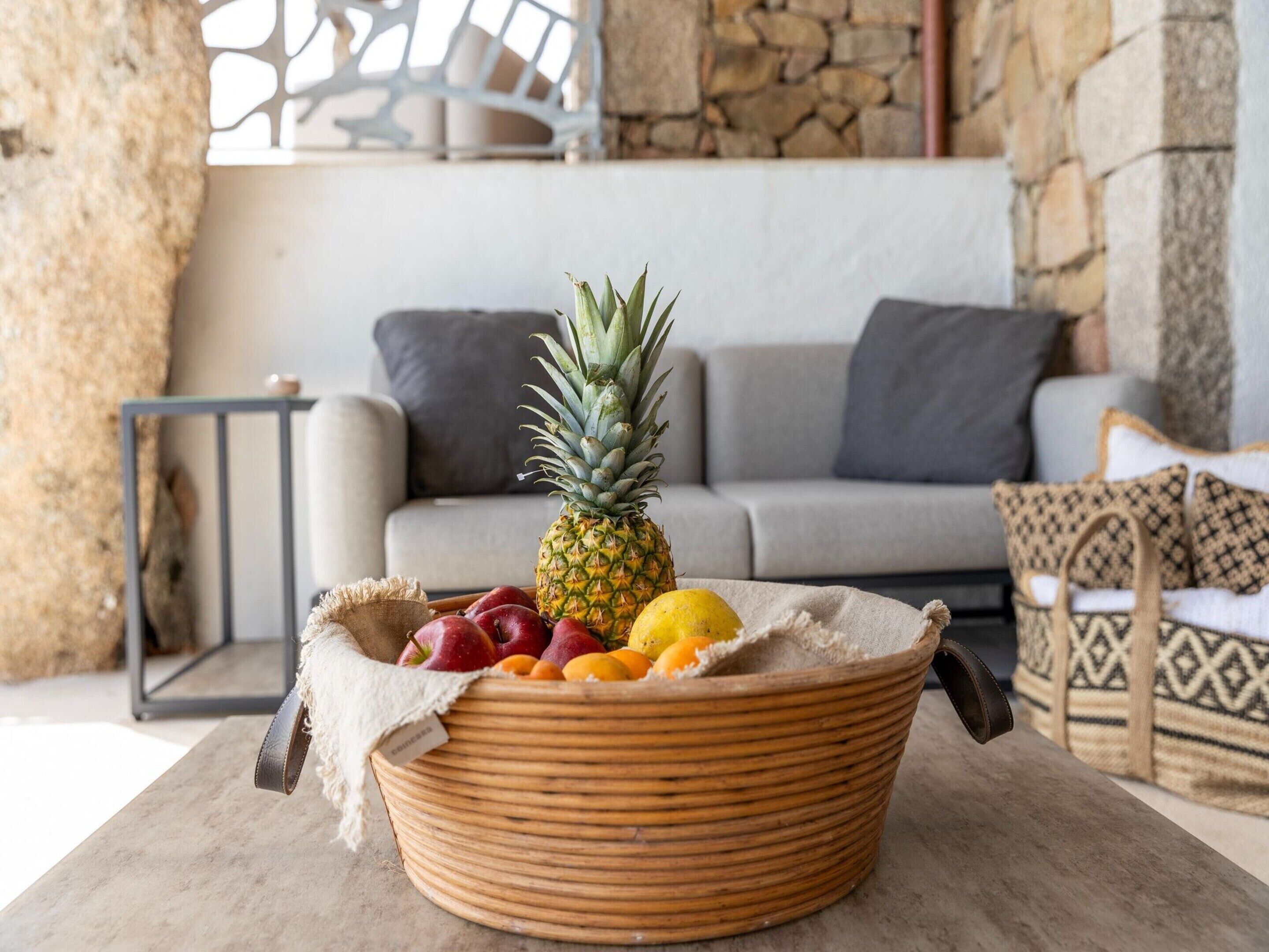 Uma cesta de vime com frutas sobre uma mesa numa sala de estar com parede de pedra e um sofá cinzento.