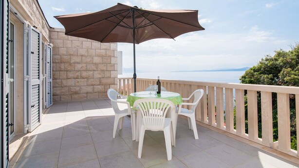 Uma varanda com mesa e cadeiras, um grande guarda-sol e vista para o mar.