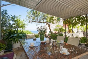Uma área de refeições ao ar livre coberta, com uma mesa posta para dois, rodeada por plantas em vasos e vista para o mar.
