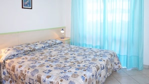Quarto com cama, mesa de cabeceira, luminária, quadro na parede e janela com cortinas azul claras.