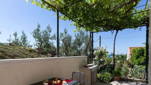 Pátio externo com mesa posta para café da manhã, cercado por cadeiras, sob uma pérgola coberta por plantas trepadeiras.