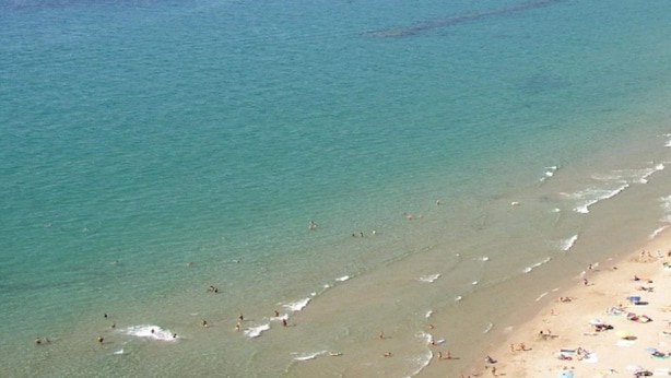 Uma praia com águas turquesa cristalinas, areal arenoso e um horizonte urbano ao longe.