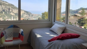Quarto com cama, poltrona e grandes janelas com vista para uma paisagem montanhosa e uma cidade.