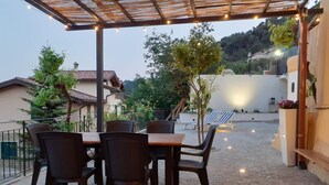 Um pátio externo com mesa e cadeiras de madeira, uma pérgula com luzes de cordão e vista da região circundante, incluindo um edifício e árvores.