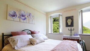 Quarto com cama, almofadas, mesa de cabeceira, candelabro e janela com vista para o verde.