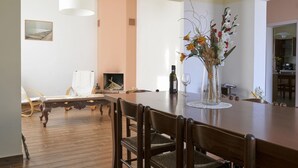 Uma área de jantar com mesa de madeira, cadeiras, um vaso com flores, uma garrafa de vinho e um quadro na parede.