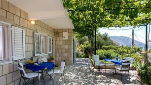 Um alpendre coberto com piso de pedra, cadeiras brancas, uma mesa com toalha azul e vista para vegetação e montanhas ao fundo.