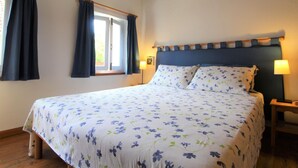 Quarto com cama, vigas de madeira no teto, janela com cortinas e abajur.