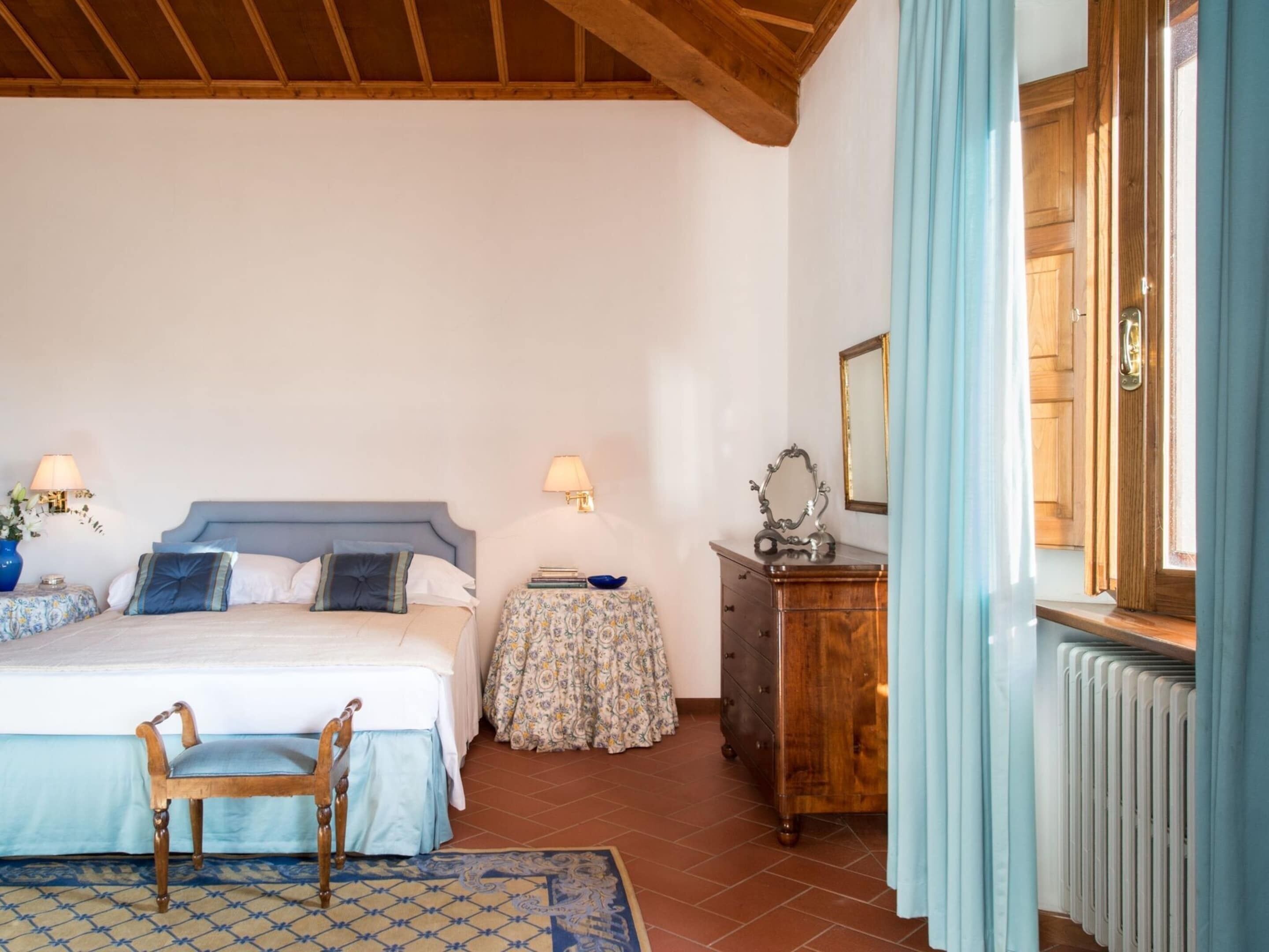 Quarto com cama, mesas de cabeceira, penteadeira, uma cadeira e janela com cortinas.