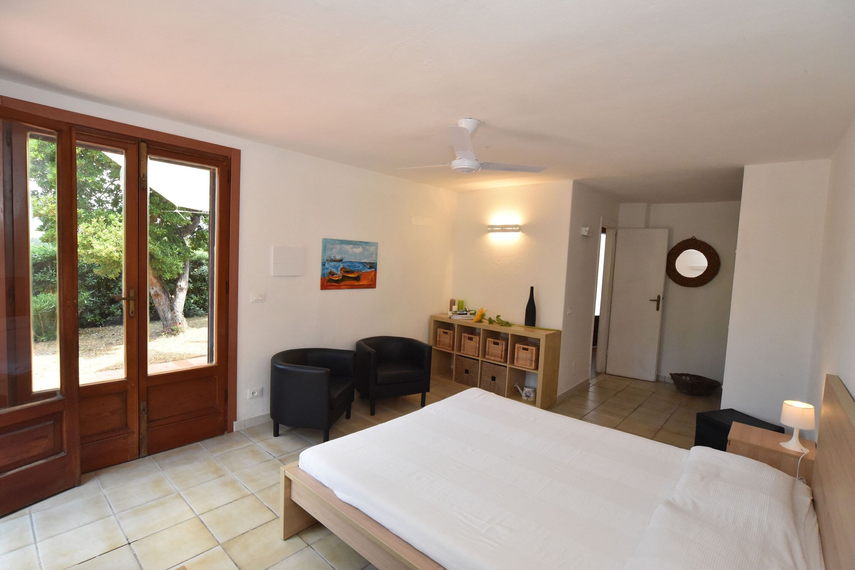 Quarto com cama, escrivaninha, duas cadeiras, ventilador de teto e vista para o exterior através de uma porta deslizante.