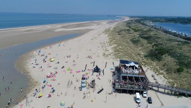Uma praia com um pavilhão e palco para eventos, rodeada por areia e dunas.
