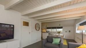 Uma sala de estar moderna com televisão de tela plana, sofás cinzas, um espaço de refeições e uma cozinha ao fundo.