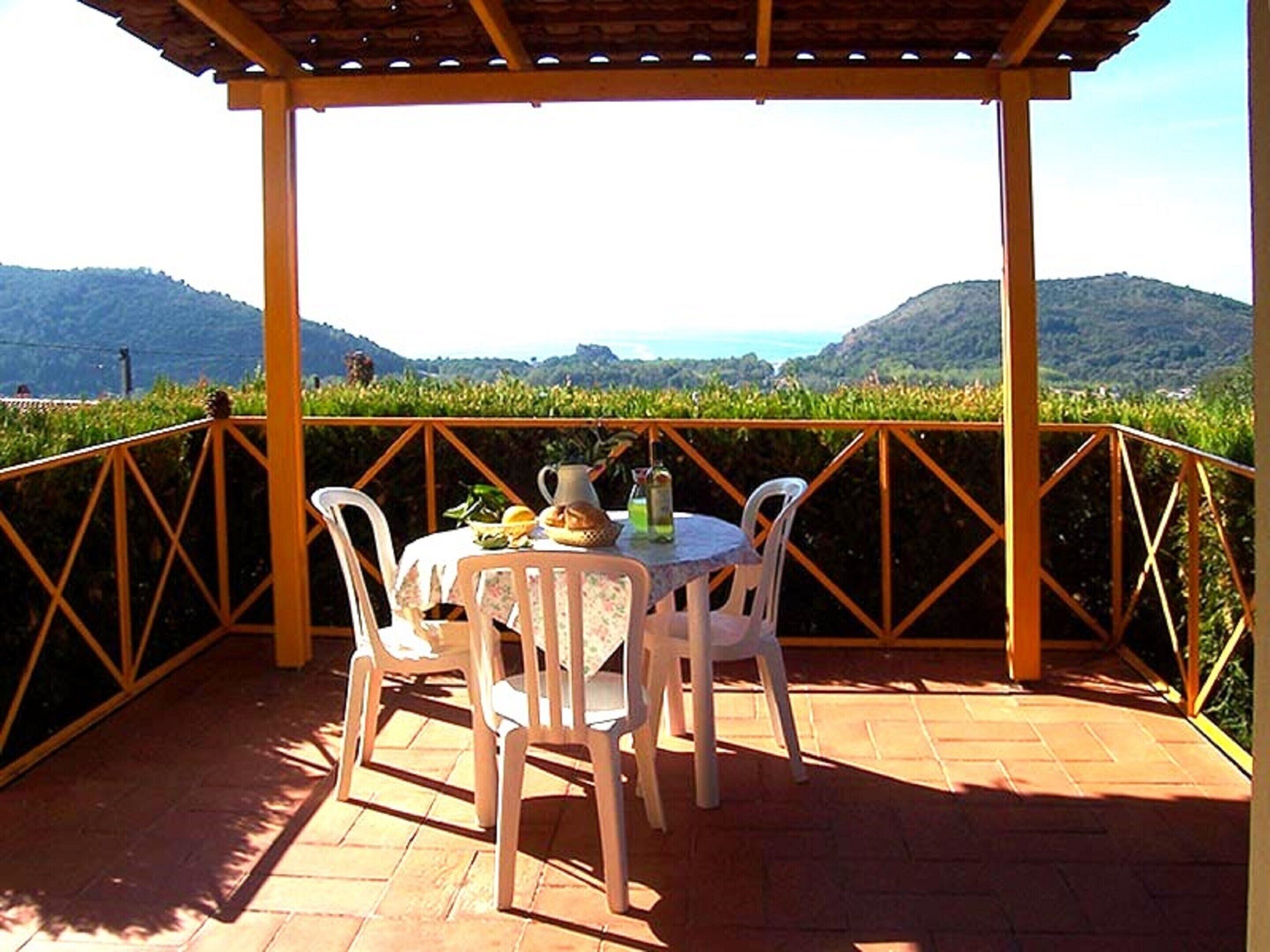 Uma área de refeições ao ar livre coberta, com mesa e cadeiras, rodeada por um guarda-corpo de metal, com vista para um vinhedo e colinas.
