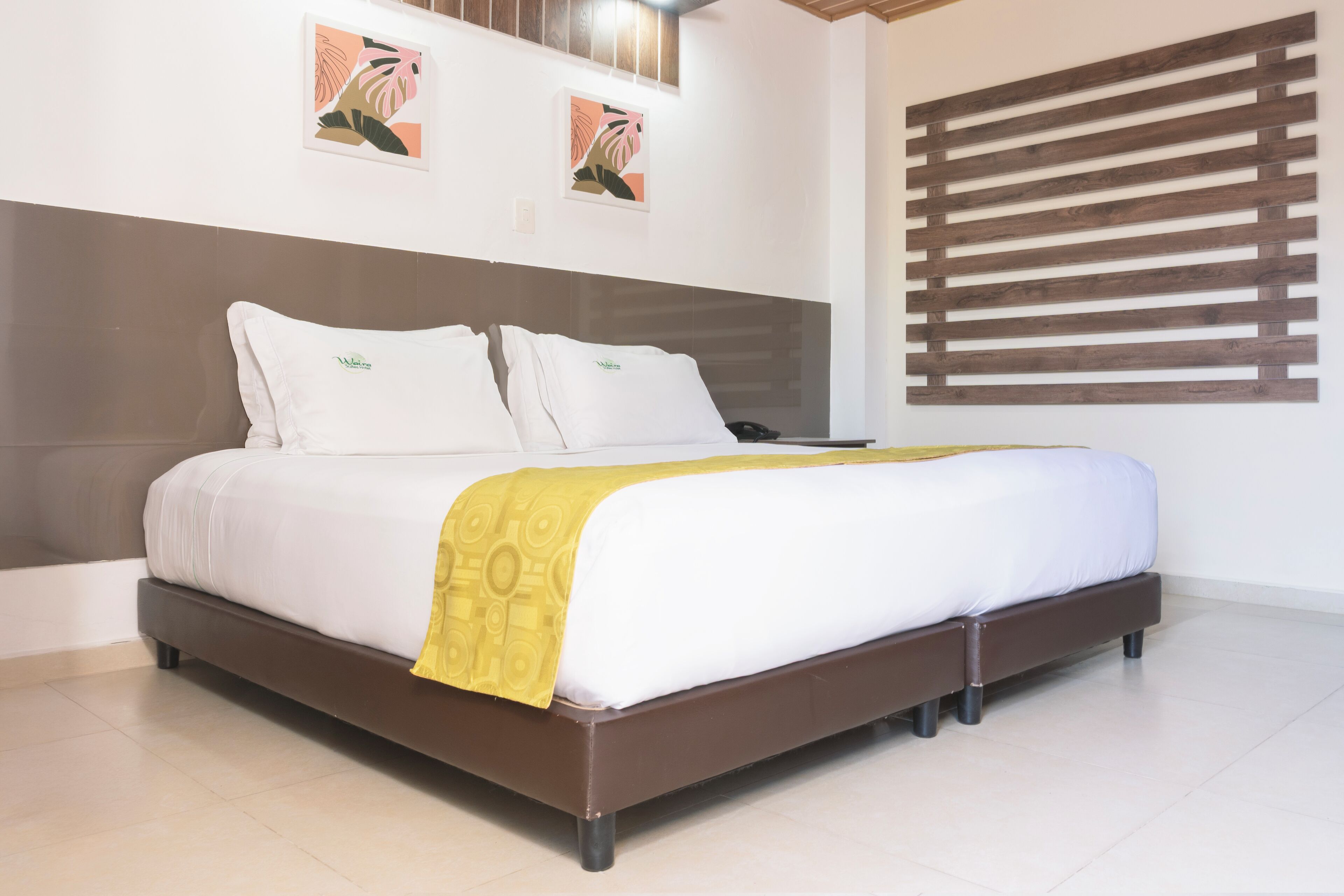 Uma cama bem arrumada com lençóis brancos e um cobertor amarelo, uma cabeceira marrom e duas obras de arte penduradas na parede.