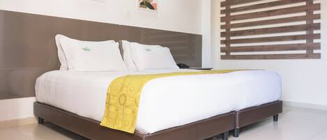 Uma cama bem arrumada com lençóis brancos e um cobertor amarelo, uma cabeceira marrom e duas obras de arte penduradas na parede.