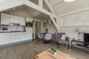 Uma cozinha moderna com armários brancos, uma área de jantar com mesa e cadeiras redondas, um espaço de estar acolhedor com sofá e um rack para televisão, e uma escada que leva a um andar superior.