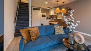 Uma sala de estar moderna com um sofá azul, uma escada, área de cozinha e iluminação decorativa.