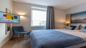 Quarto de hotel moderno com uma cama grande, uma escrivaninha e uma cadeira. Há uma janela com vista para uma ponte e um quadro na parede.