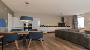 Sala de estar moderna com sofá, mesa de jantar e cadeiras. Possui área de cozinha com eletrodomésticos e uma janela com cortinas.