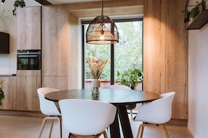 Uma cozinha moderna com armários de madeira, forno embutido, mesa de jantar com cadeiras brancas e uma janela grande com vista para o verde.