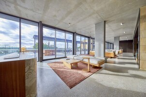 Um saguão de hotel moderno com janelas amplas, um balcão de recepção, áreas de estar e vista para o exterior.