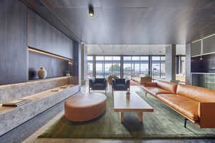 Uma sala de estar moderna com um sofá de couro, um banquinho redondo e uma mesa de centro. Há duas poltronas e uma estante com objetos decorativos. Grandes janelas oferecem vista para o exterior.