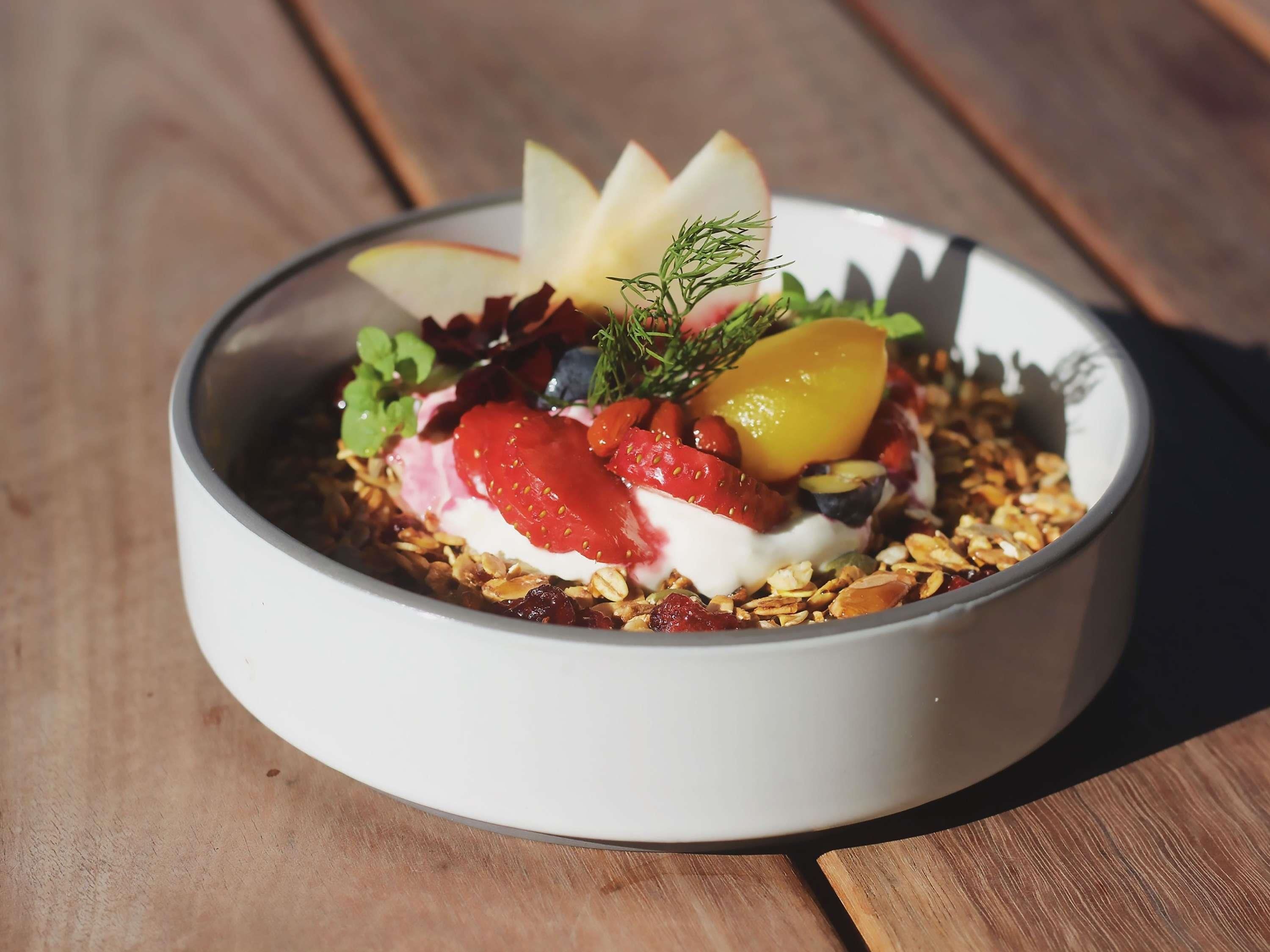 Uma tigela de comida com diversas frutas, nozes e uma colherada de creme sobre uma mesa de madeira.