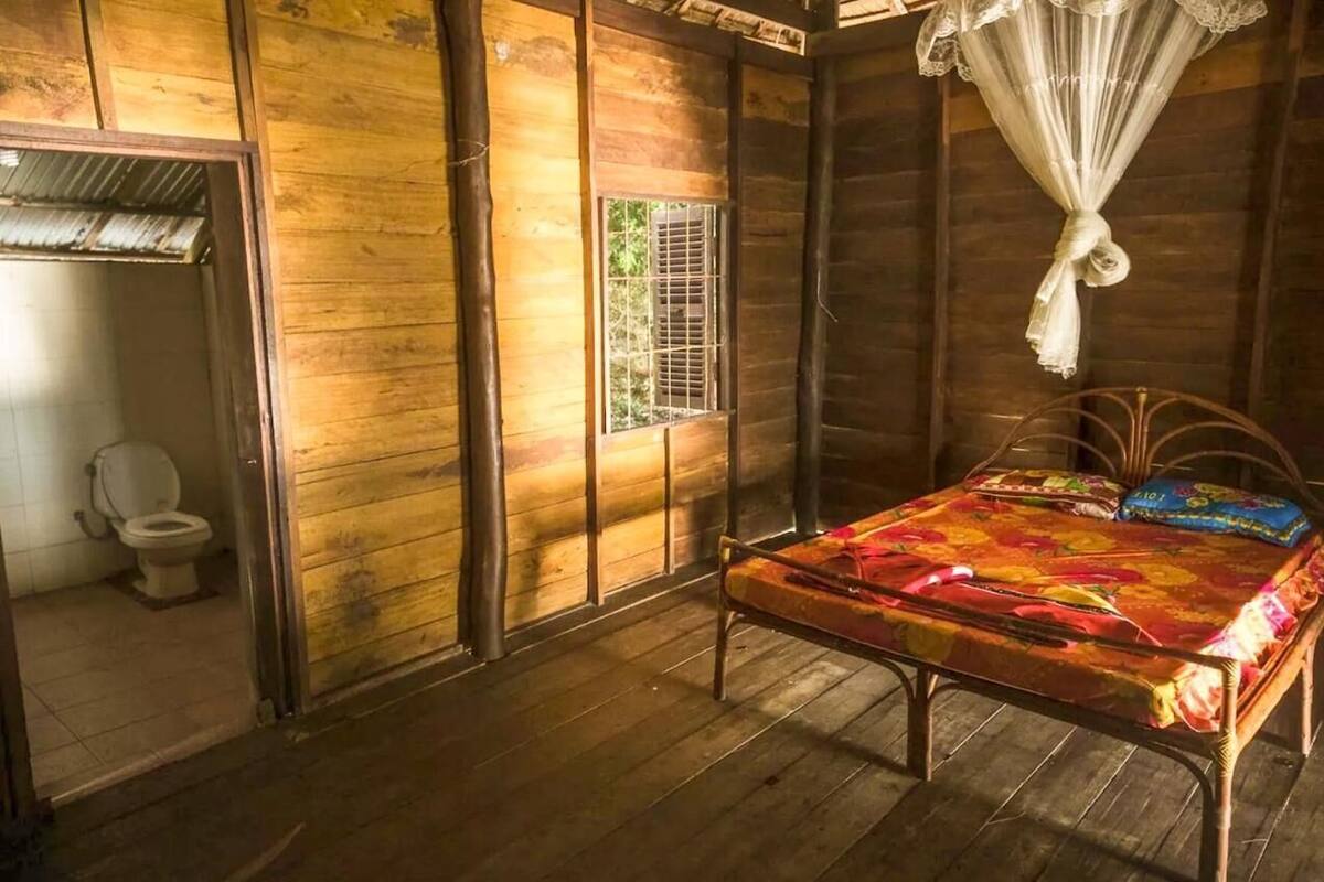 Um quarto de madeira com uma cama, uma janela com grades e um vaso sanitário.