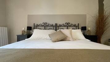Cama bem arrumada com cabeceira, duas mesas de cabeceira com abajures e uma planta alta ao lado da cama.