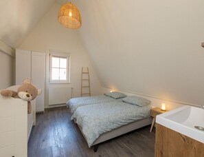 Quarto com teto inclinado, cama com manta estampada, abajur, escada de madeira, urso de pelúcia e janela com persianas.