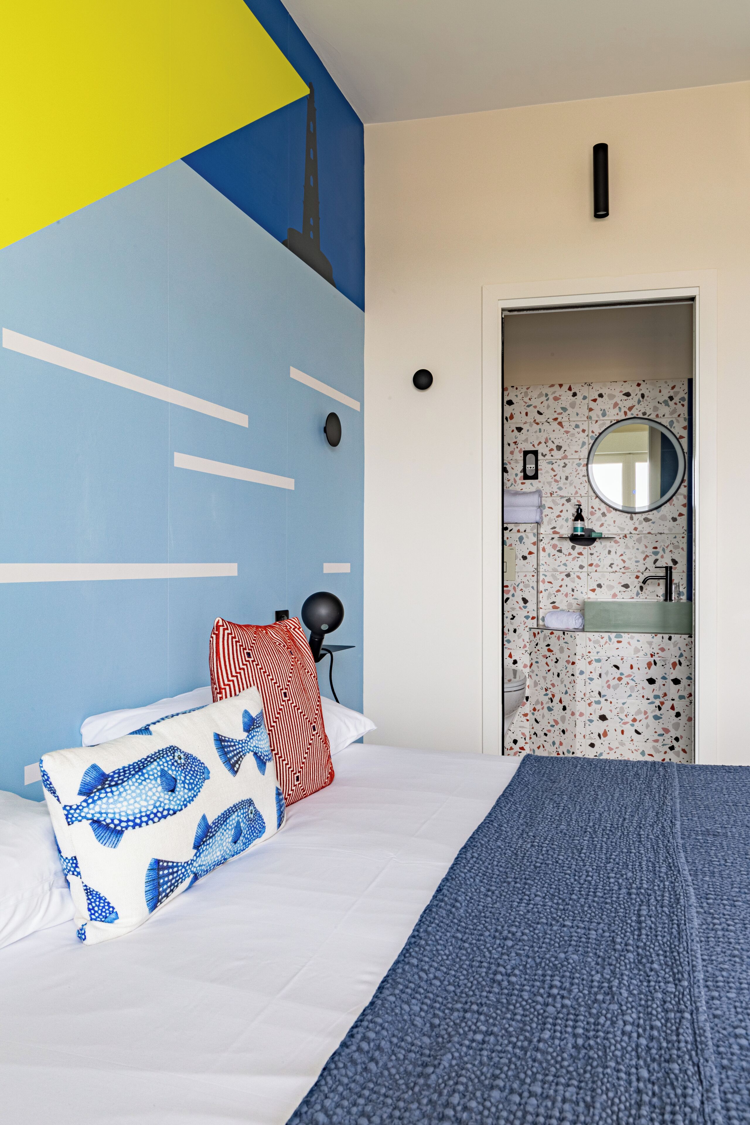 Quarto com cama, travesseiro estampado em azul e branco, travesseiro vermelho e branco, manta azul, parede com design geométrico, espelho e banheiro com parede azulejada e pia.