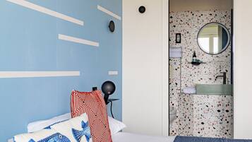 Quarto com cama, travesseiro estampado em azul e branco, travesseiro vermelho e branco, manta azul, parede com design geométrico, espelho e banheiro com parede azulejada e pia.