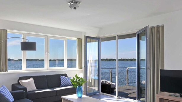 Uma sala de estar moderna com sofá, mesa de centro e rack para TV, grandes janelas com vista para um lago e porta de vidro deslizante para um deck externo.