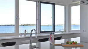 Cozinha moderna com uma ilha grande, pia de aço inoxidável e três janelas com vista para a água.