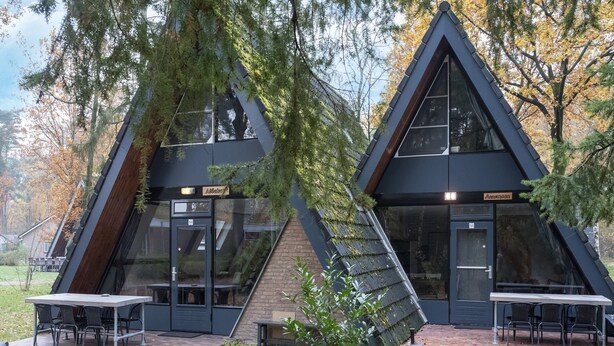 Dois edifícios modernos, com formato triangular e fachadas de vidro, área externa com mesas e cadeiras, e uma pequena árvore em primeiro plano.