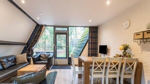 Uma sala de estar moderna com sofá, mesa de centro e área de jantar com cadeiras e uma mesa de madeira. Uma grande janela oferece vista para árvores.