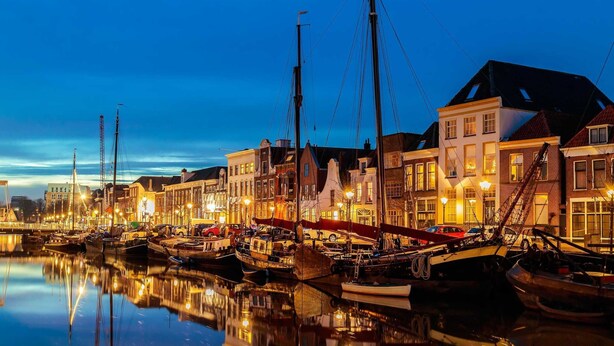 Uma marina à beira-mar com barcos atracados, edifícios com iluminação acolhedora e um céu claro.