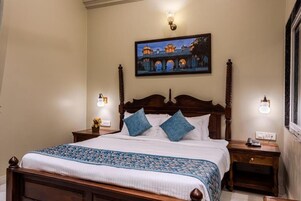 Cama bem arrumada com colcha estampada, duas almofadas e cabeceira de madeira. Há duas mesas de cabeceira com abajures, um quadro na parede e uma janela à direita.