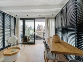 Uma sala de jantar moderna com mesa de madeira, cadeiras brancas e vista para um canal e casas através de amplas janelas.