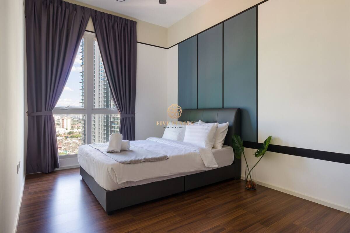 Quarto de hotel moderno com uma cama grande, cabeceira escura e vista da cidade através de amplas janelas.