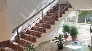 Escada com corrimãos de metal que leva a um andar superior, cercada por plantas em vasos e um tapete vermelho sobre piso de azulejos.