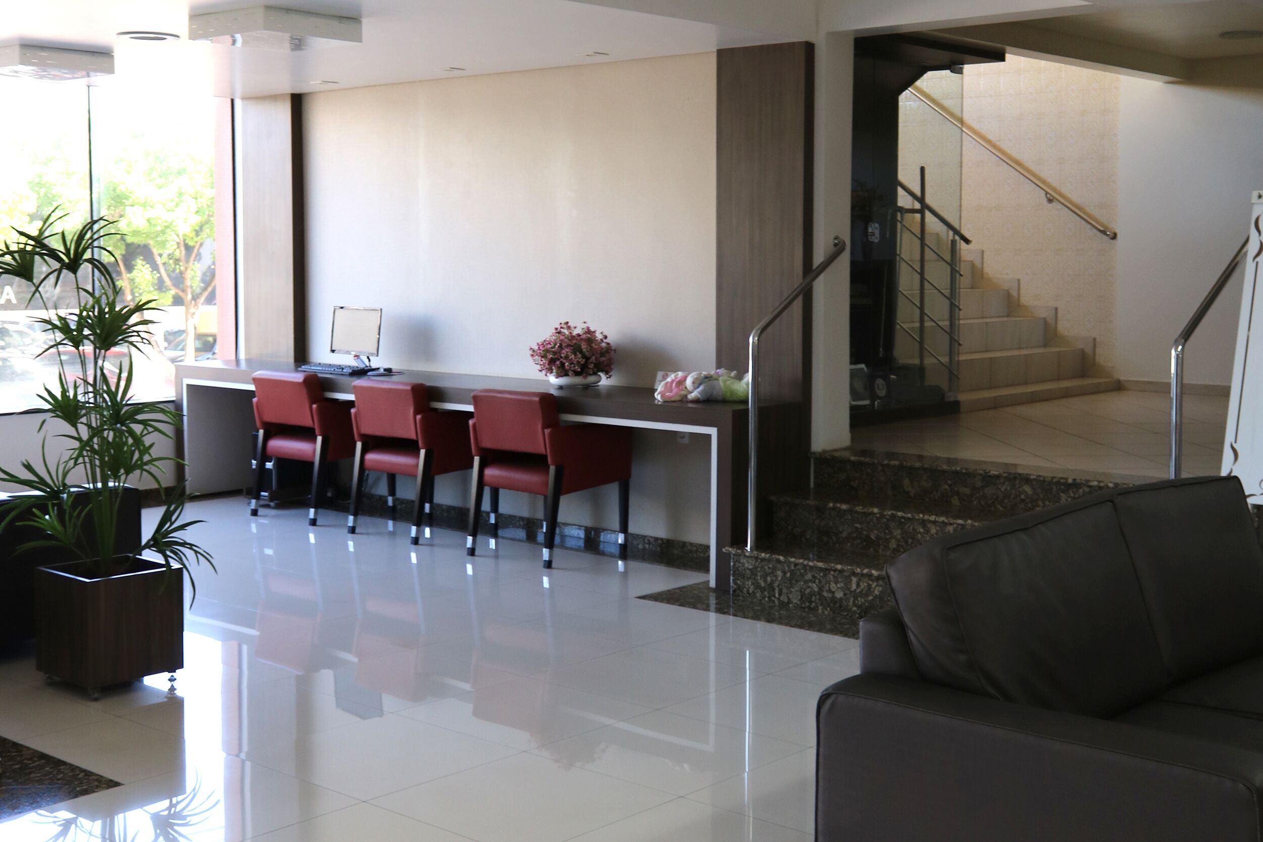 Um lobby moderno com uma mesa de recepção, cadeiras vermelhas, uma planta em vaso e uma escada.