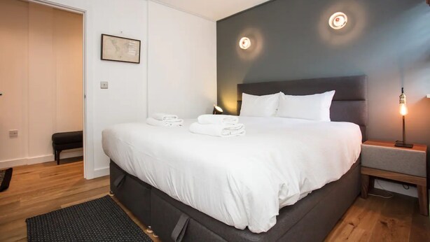 Quarto moderno com uma cama grande, mesa de cabeceira com luminária, um banquinho e um quadro na parede.