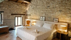 Quarto com parede de pedra, cama com roupa de cama branca, duas luminárias de cabeceira e um banco de madeira.
