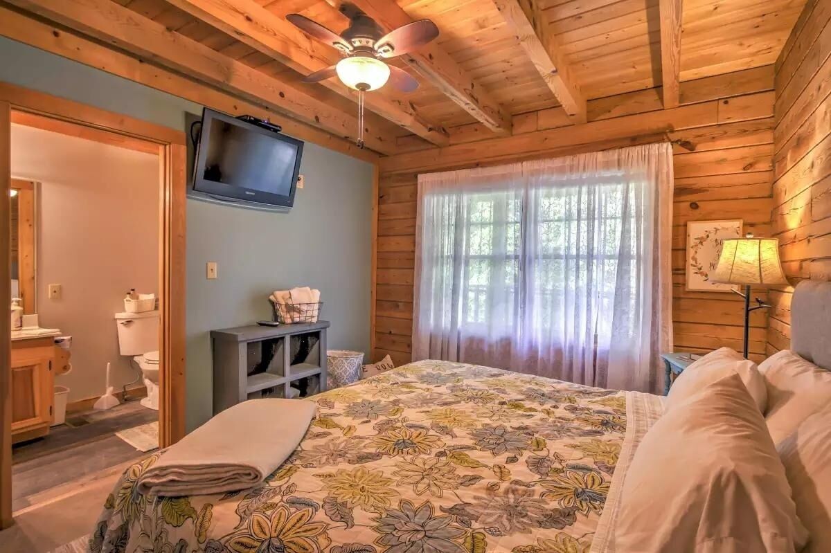 Quarto de cabana de madeira com cama, televisão, ventilador de teto e janela com cortinas.