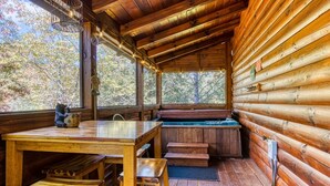 Interior de cabana de madeira com mesa, cadeiras, um banco, banheira de hidromassagem e grandes janelas com vista para árvores.