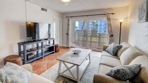 Sala de estar com televisão, sofá, mesa de centro e varanda com mobiliário de exterior.