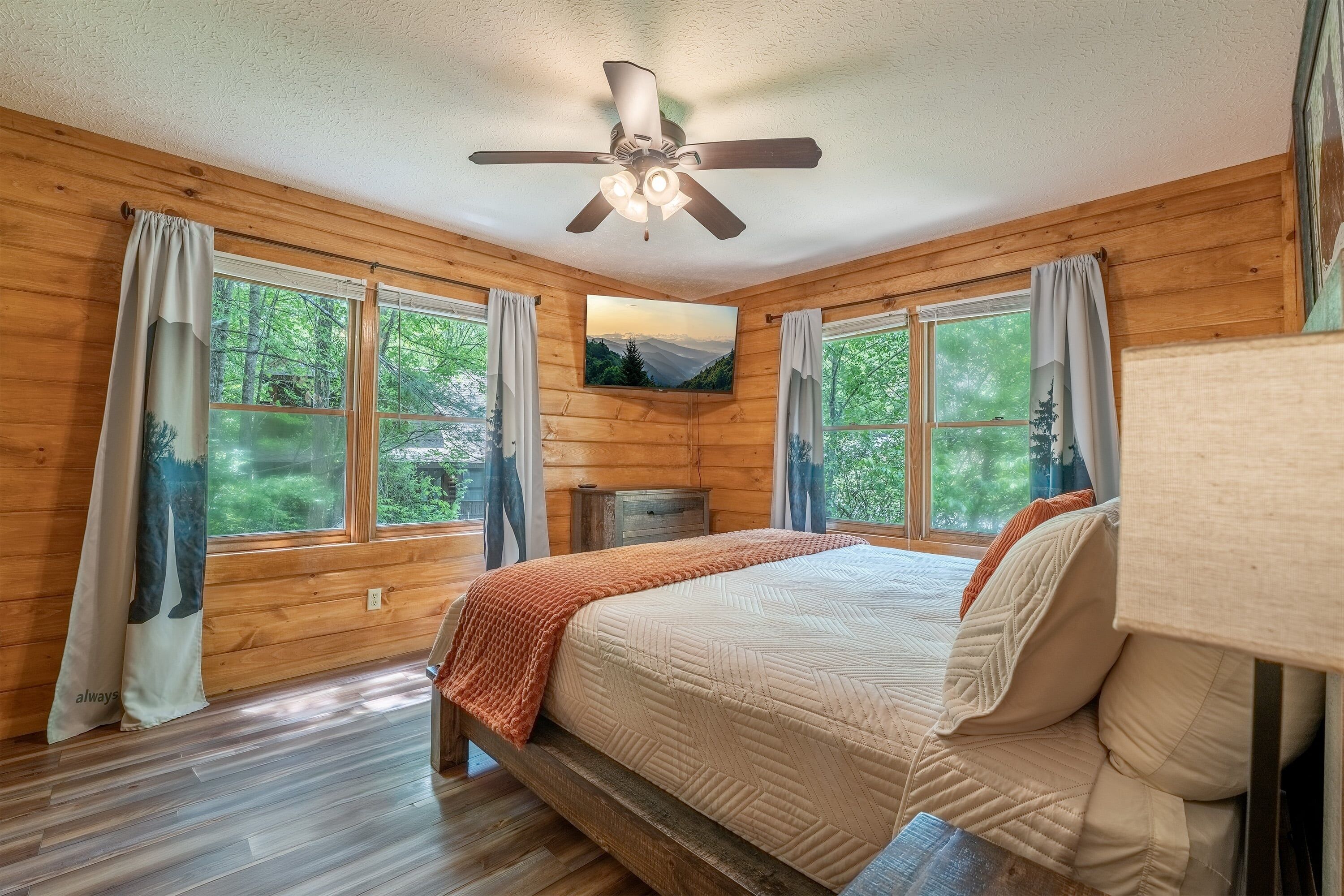 Quarto de cabana de madeira com cama, ventilador de teto e janelas amplas.